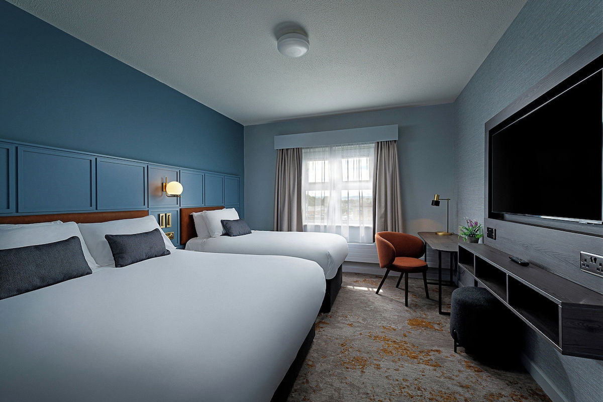 Quarto de hotel com duas camas, uma escrivaninha, televisão e janela com cortinas.