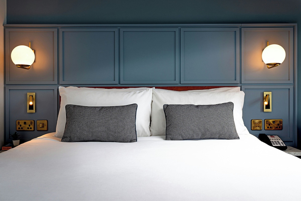 Quarto de hotel com cama grande, duas luminárias fixadas na parede e mesa de cabeceira com telefone.