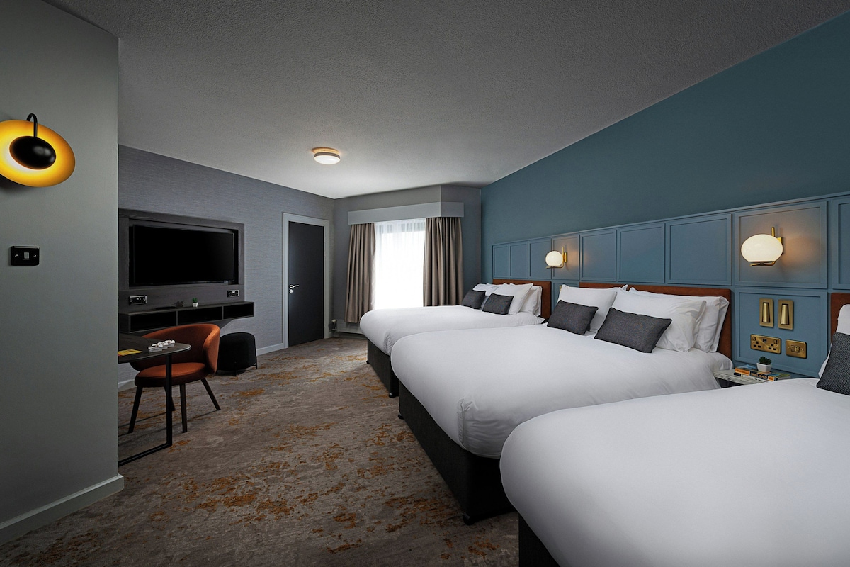 Quarto de hotel com duas camas, uma escrivaninha com cadeira, televisão e janela com cortinas.