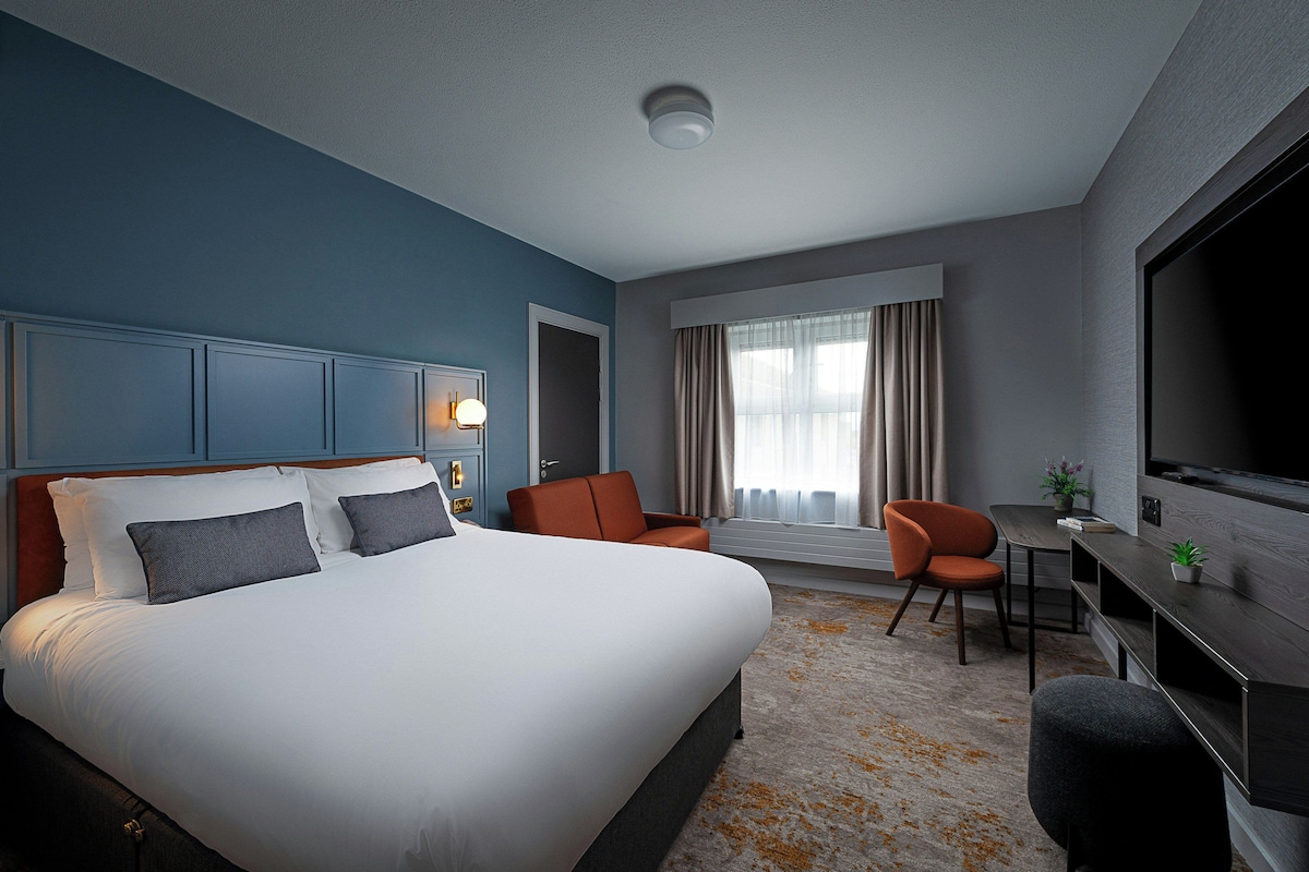 Quarto de hotel com uma cama grande, escrivaninha, cadeira e televisão.