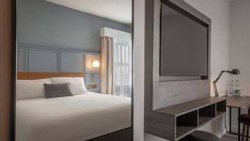 Quarto de hotel com cama, televisão na parede, escrivaninha com luminária e uma cadeira.