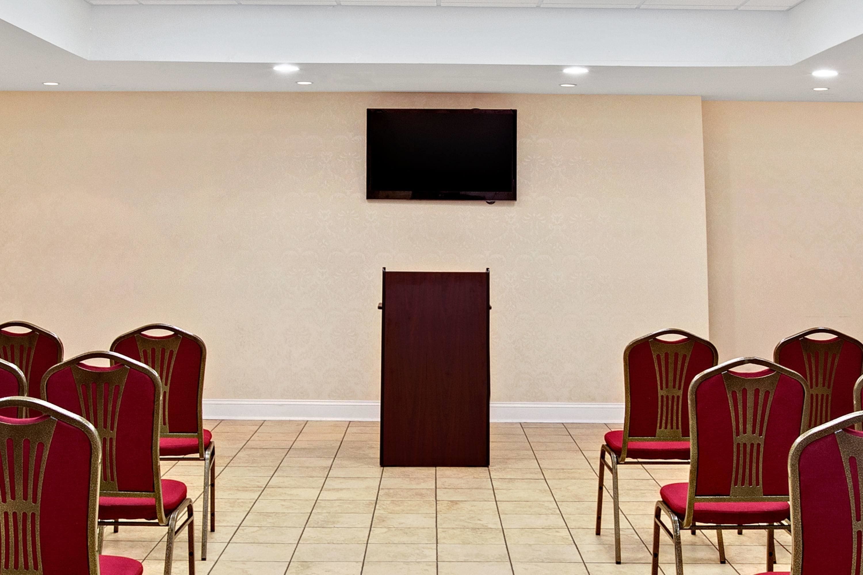 Sala de conferências com cadeiras vermelhas, um púlpito de madeira e uma TV de tela plana na parede.
