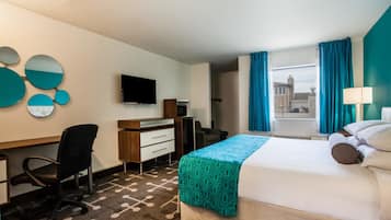 Quarto de hotel com cama, escrivaninha, cadeira, televisão e janela com cortinas.