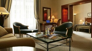Quarto de hotel com sofá, duas poltronas, mesa de centro de vidro, escrivaninha com um laptop e cama com cabeceira vermelha.
