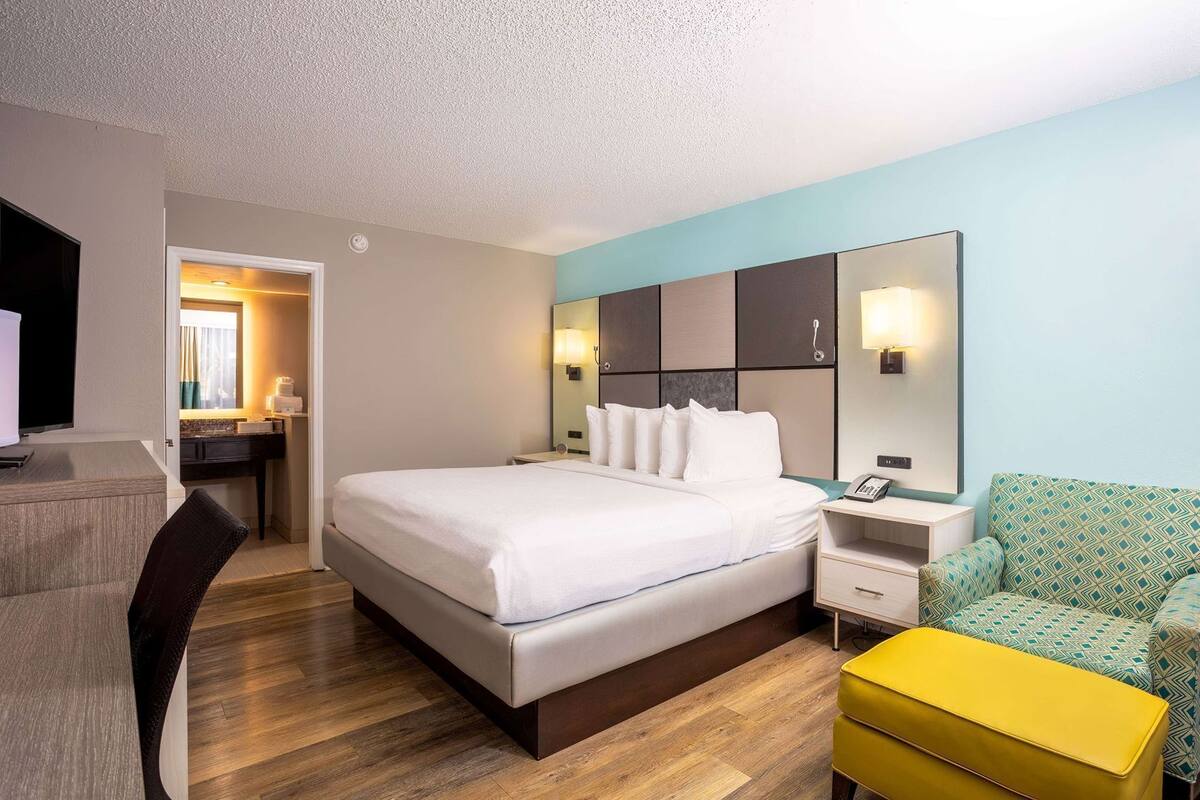 Quarto de hotel com cama, mesa de cabeceira, espelho e um banquinho amarelo.