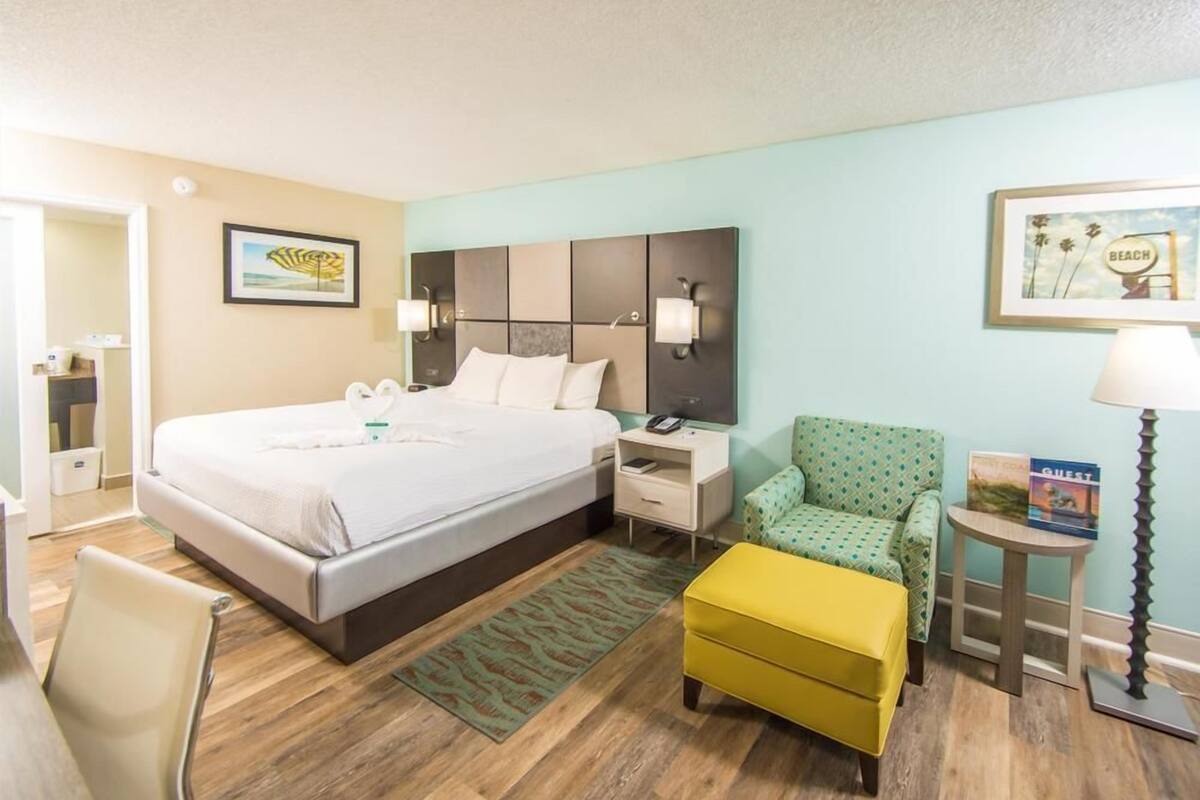 Quarto de hotel com cama, mesa de cabeceira, cadeira, pequena mesa e abajur.