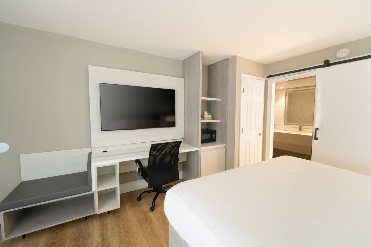 Quarto de hotel com uma cama, uma escrivaninha com cadeira, televisão, guarda-roupa e banheiro visível por uma porta aberta.