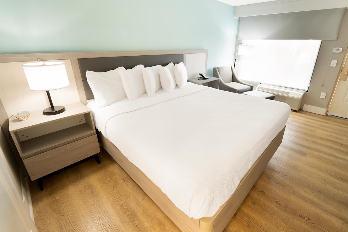 Quarto de hotel moderno com uma cama grande, mesa de cabeceira com luminária, uma cadeira e uma janela ampla.