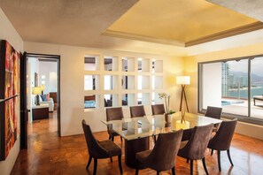 Quarto de hotel moderno com mesa de jantar de vidro, assentos confortáveis, parede decorativa e uma grande janela com vista para a cidade.