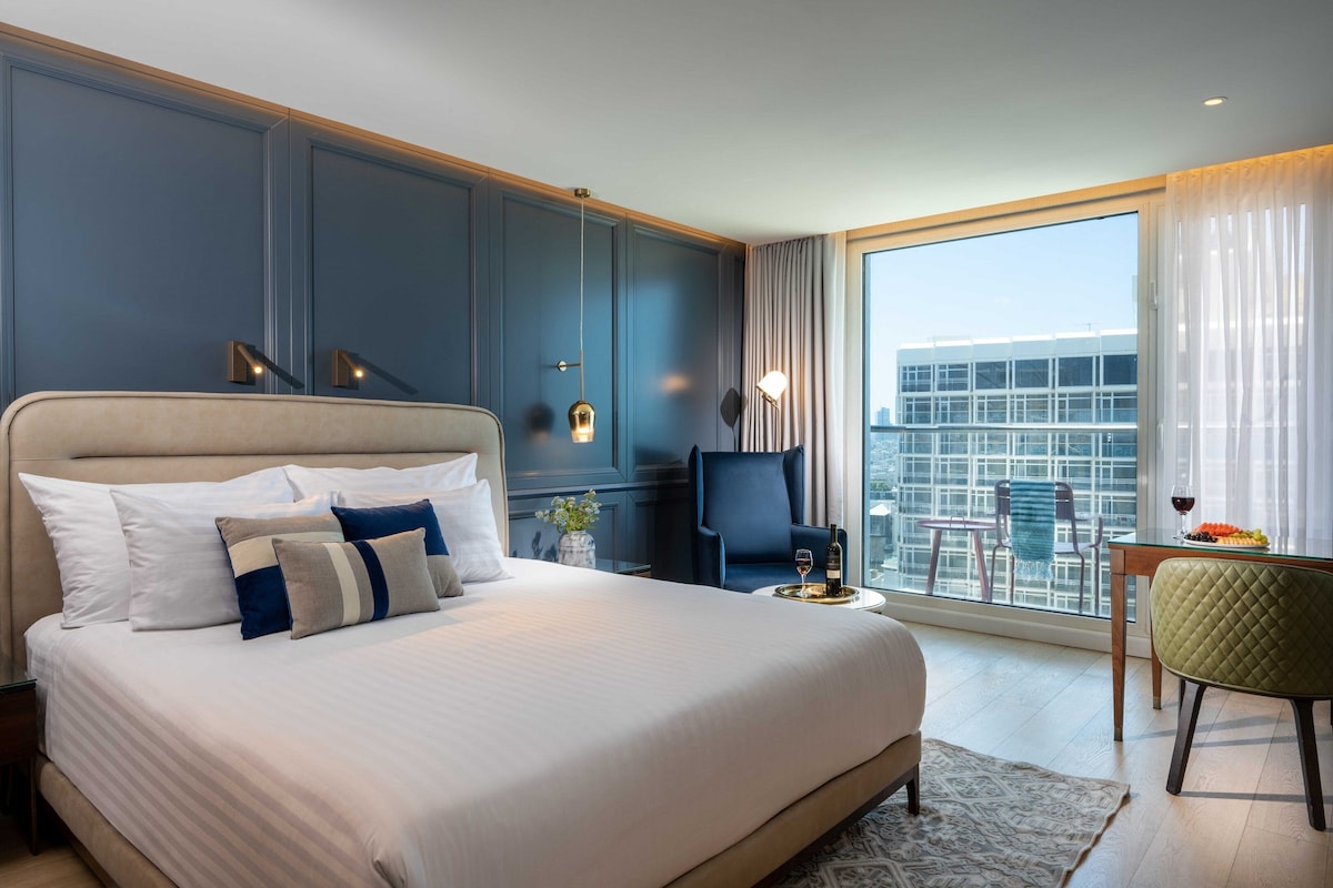 Quarto de hotel com uma cama grande, vista da cidade, área de refeições e varanda com mesa e cadeiras.