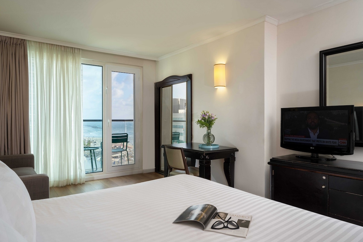 Quarto de hotel com uma cama grande, uma escrivaninha com um laptop, uma televisão e vista para o oceano pelas janelas.