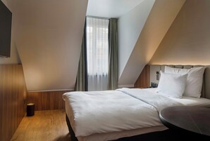 Quarto de hotel moderno com uma cama, mesa de cabeceira, luminária e janela com cortinas.