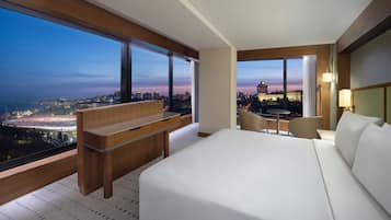 Quarto de hotel com uma cama grande, uma escrivaninha, uma cadeira e vista para a paisagem urbana ao entardecer.