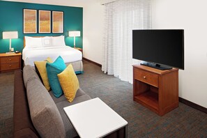 Quarto de hotel com cama, televisão sobre um suporte de madeira, sofá com almofadas, mesa pequena e mesa de cabeceira com abajur.
