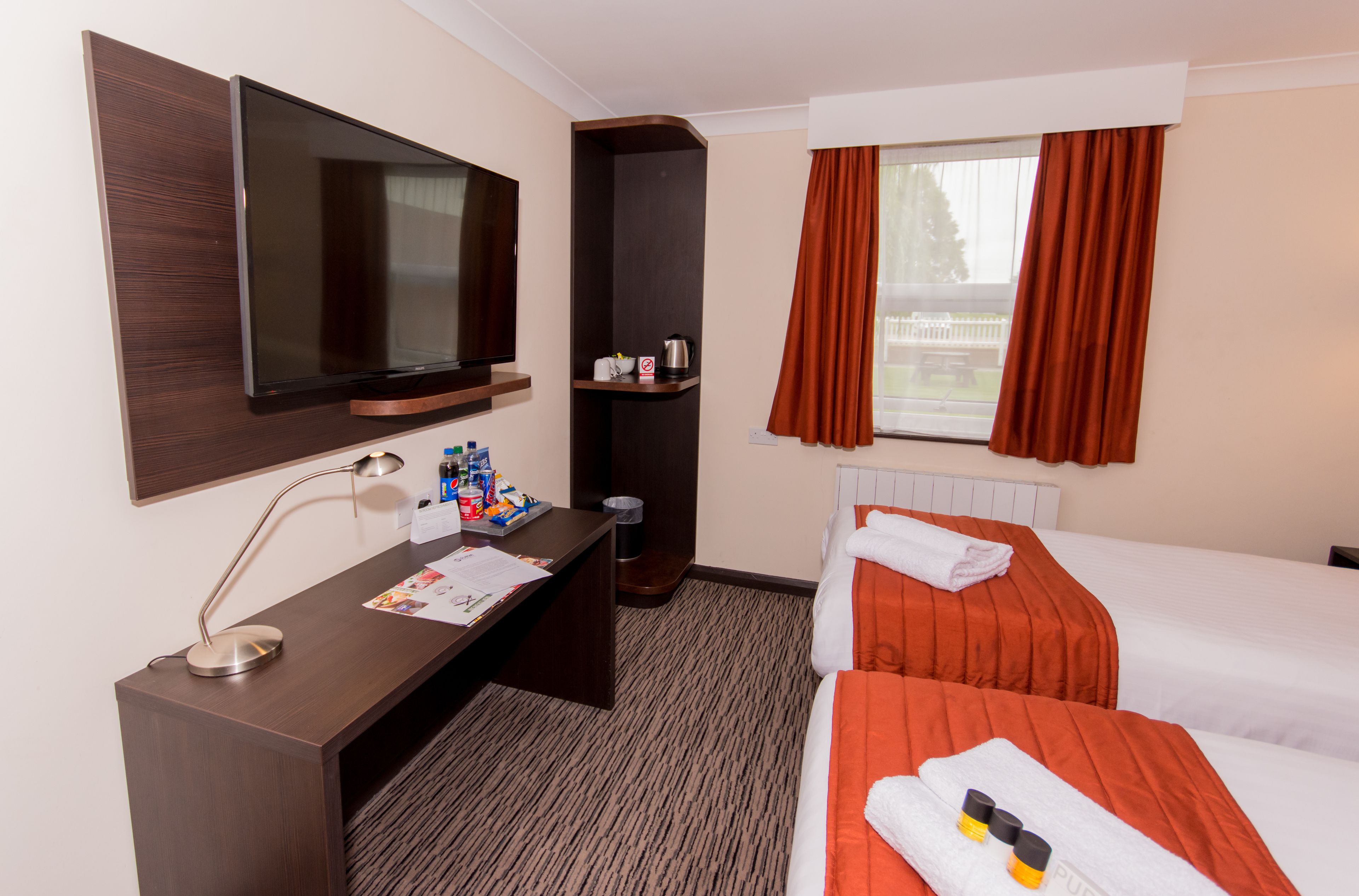 Quarto de hotel com televisão, escrivaninha, duas camas, janela com cortinas e um pequeno armário com artigos de higiene pessoal.