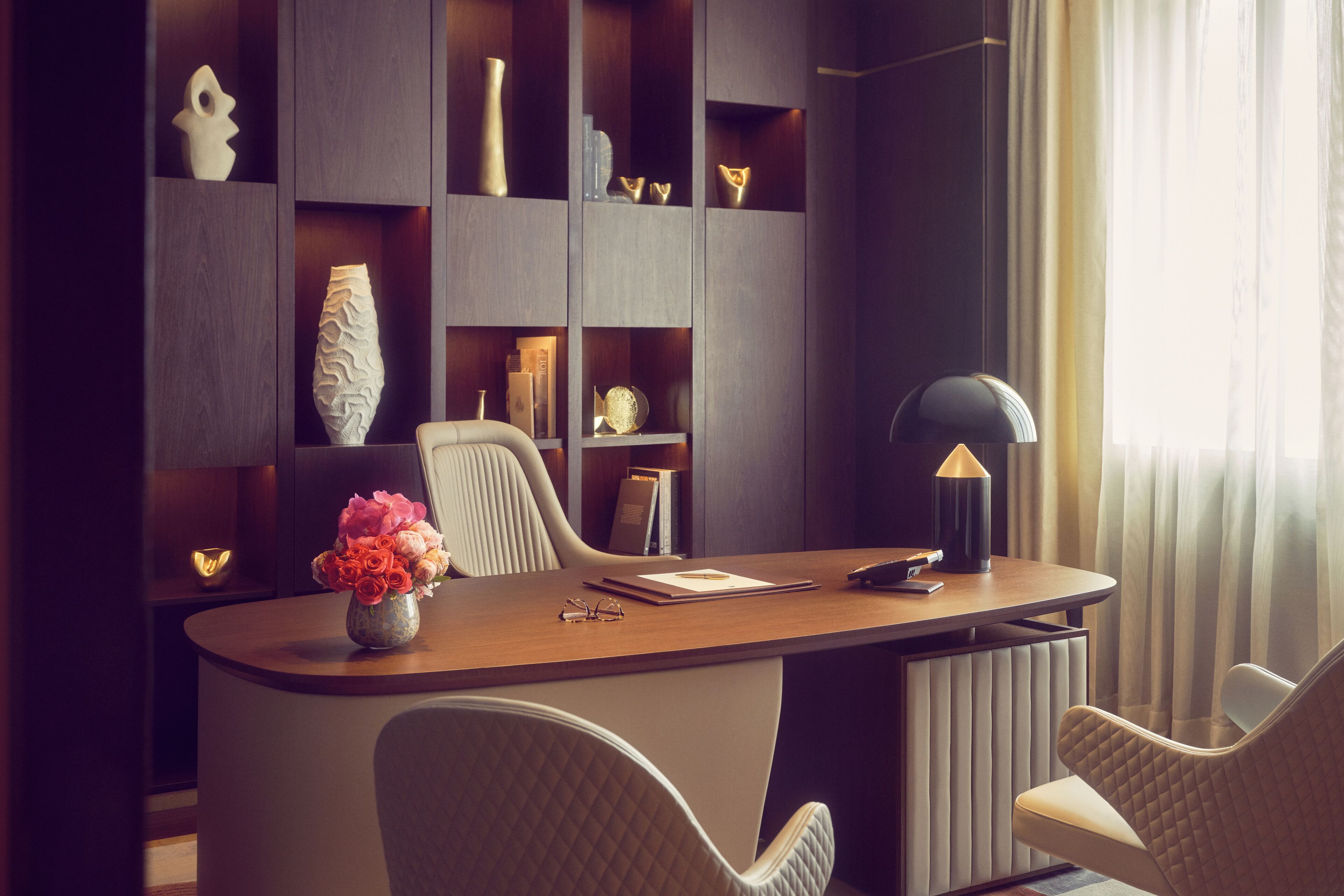 Um espaço de escritório moderno com uma mesa de madeira, duas cadeiras, um vaso de flores e prateleiras com objetos decorativos.