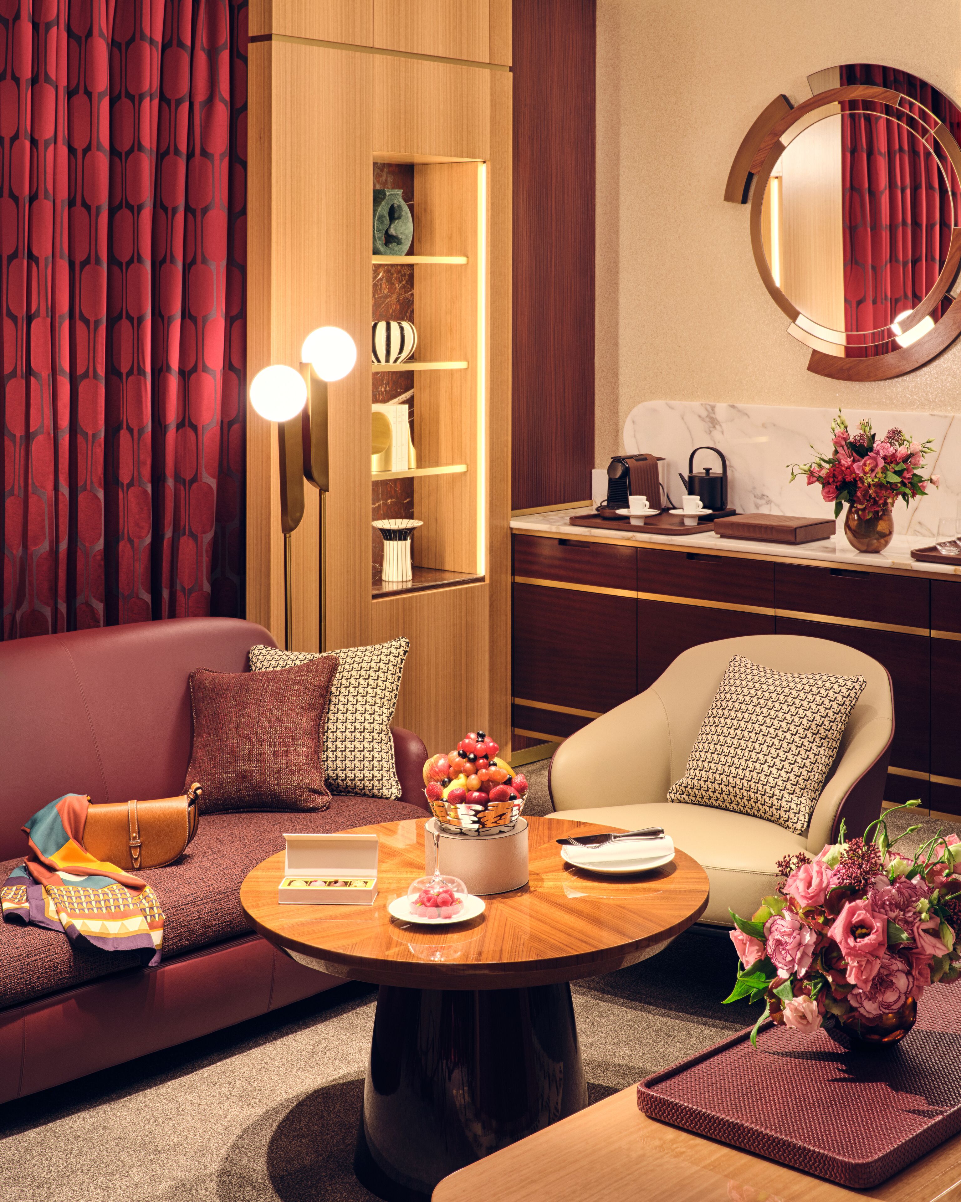 Uma sala de estar moderna com sofá, poltrona e mesa de centro. Há uma pequena mesa com um vaso de flores e uma tigela de frutas. Um espelho está pendurado na parede, e uma estante com objetos decorativos é visível.