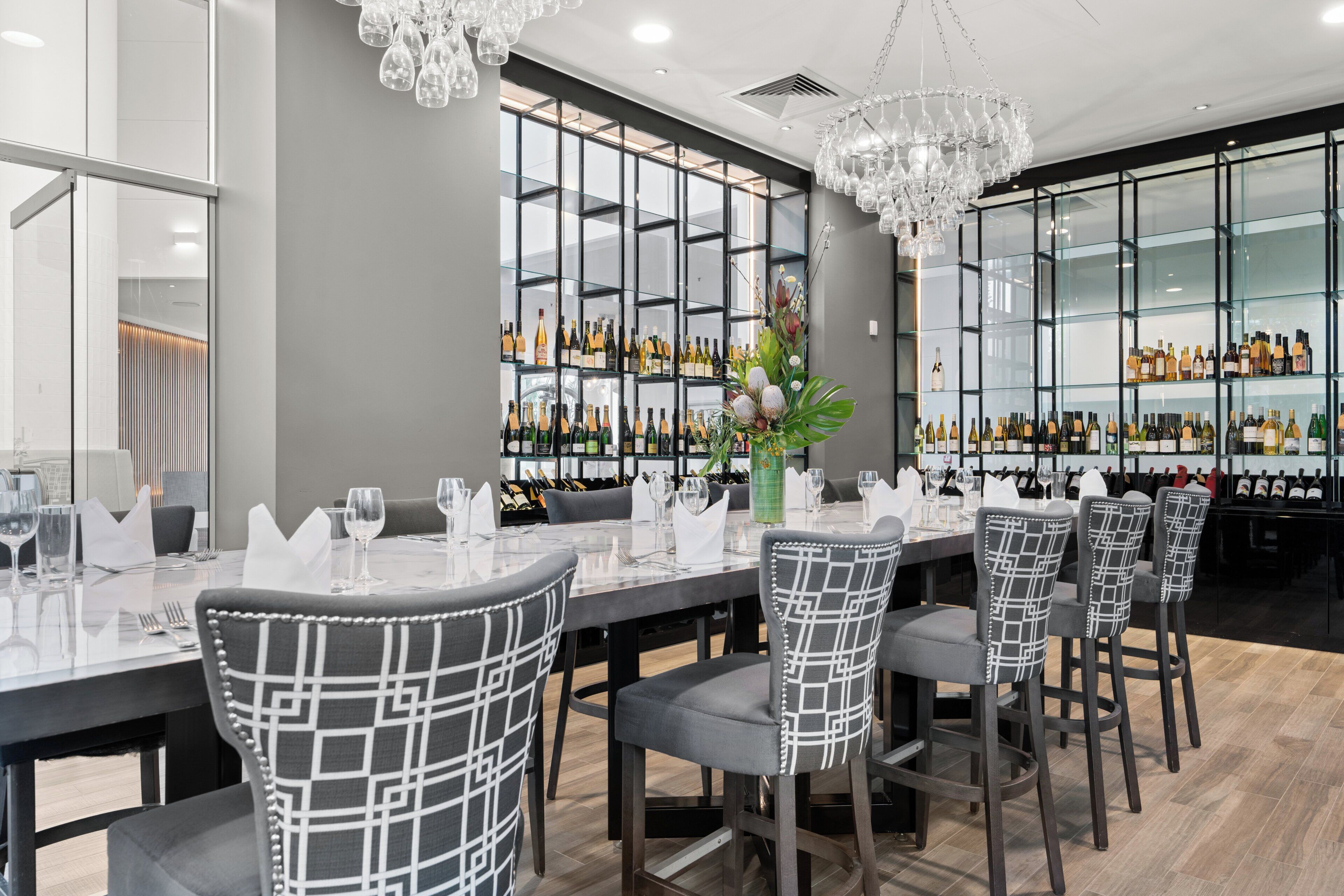 Restaurante com uma mesa comprida posta para refeição, cadeiras com estofados estampados e uma adega ao fundo.