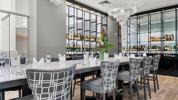 Restaurante com uma mesa comprida posta para refeição, cadeiras com estofados estampados e uma adega ao fundo.