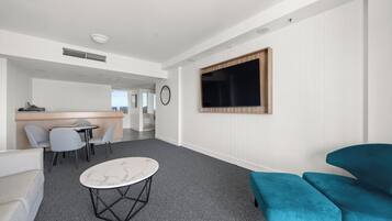 Quarto de hotel moderno com carpete cinza, mesa redonda de mármore, sofás verde-água, televisão presa a uma parede branca e janela com moldura de madeira.