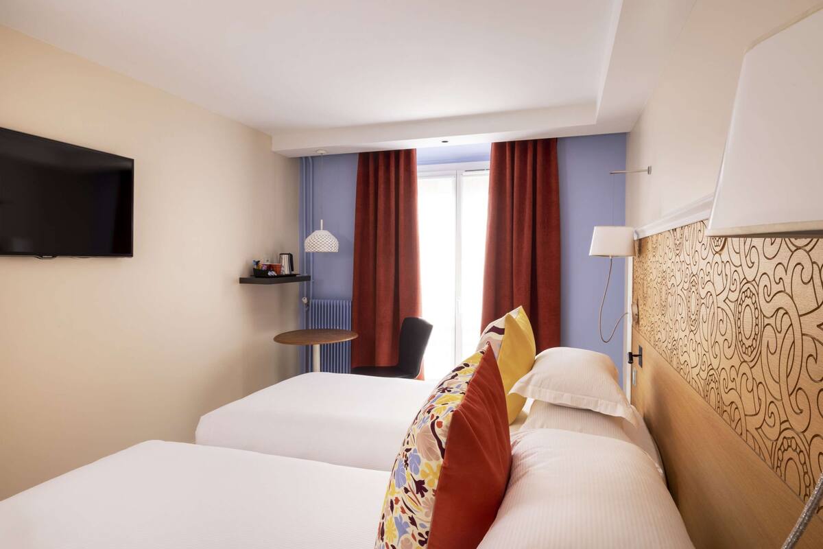 Quarto de hotel com duas camas, uma TV presa à parede, uma mesinha com candeeiro e vista exterior através de amplas janelas.