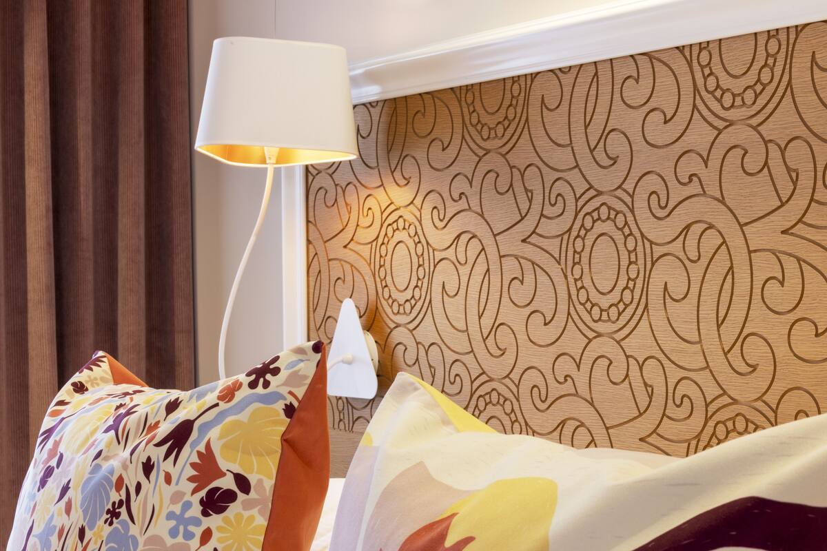 Quarto de hotel com papel de parede estampado, cama com almofadas coloridas e abajur.