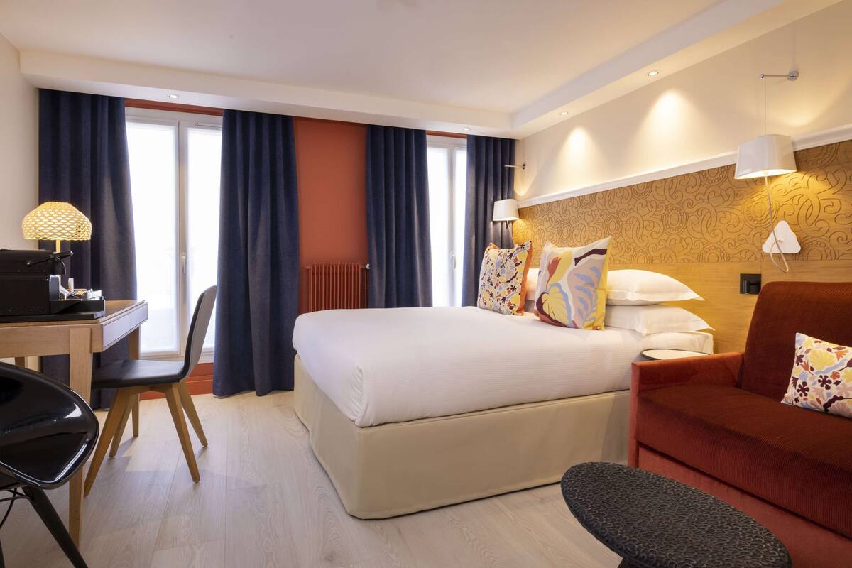 Quarto de hotel com cama, escrivaninha, cadeira, sofá e janela com cortinas.