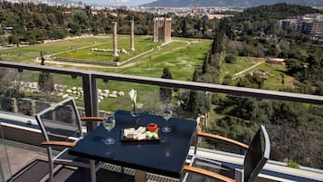 Varanda com mesa posta para dois, com vista para um parque com colunas antigas e um horizonte urbano ao fundo.