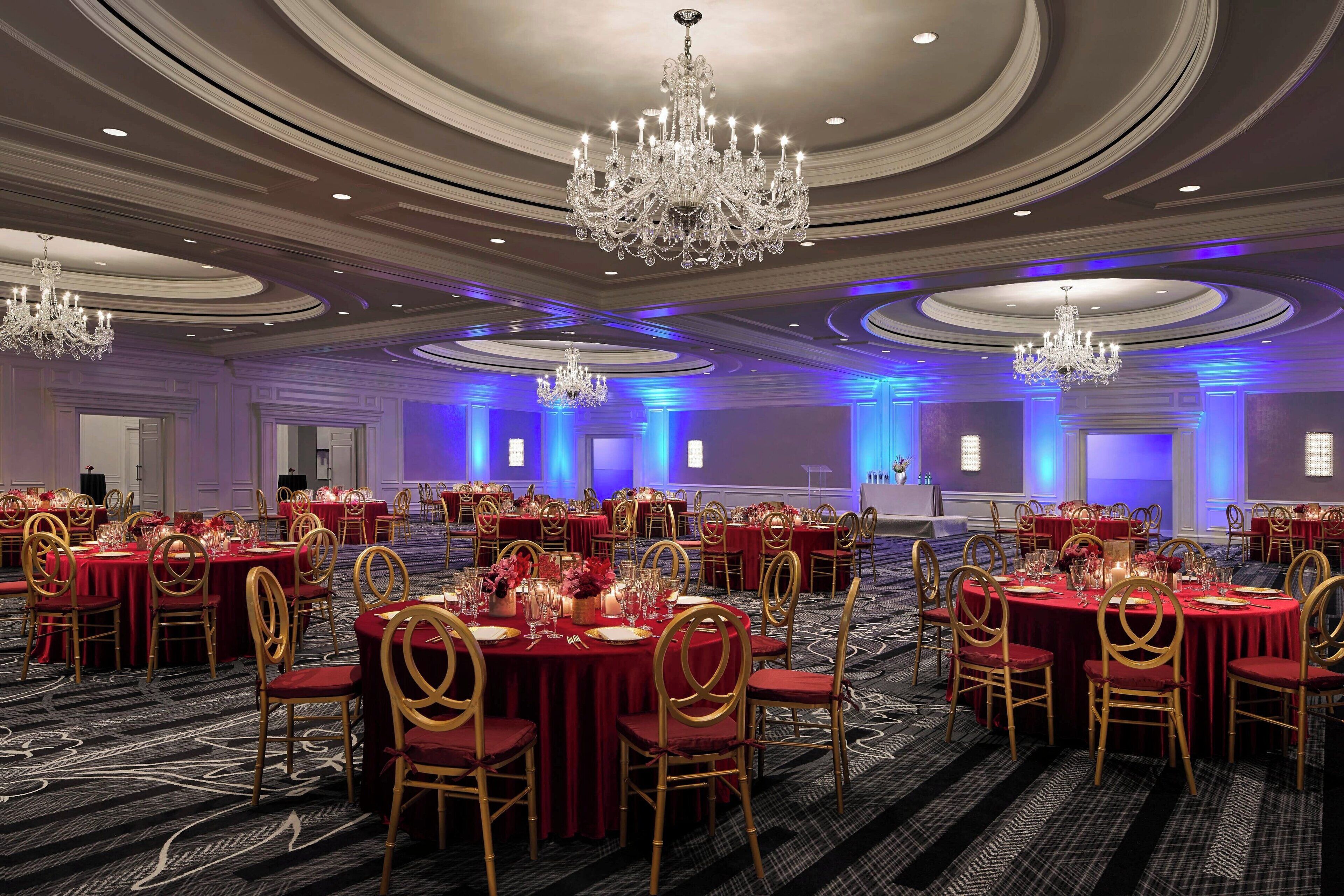 Uma sala de banquetes com mesas redondas, cadeiras douradas e lustres.