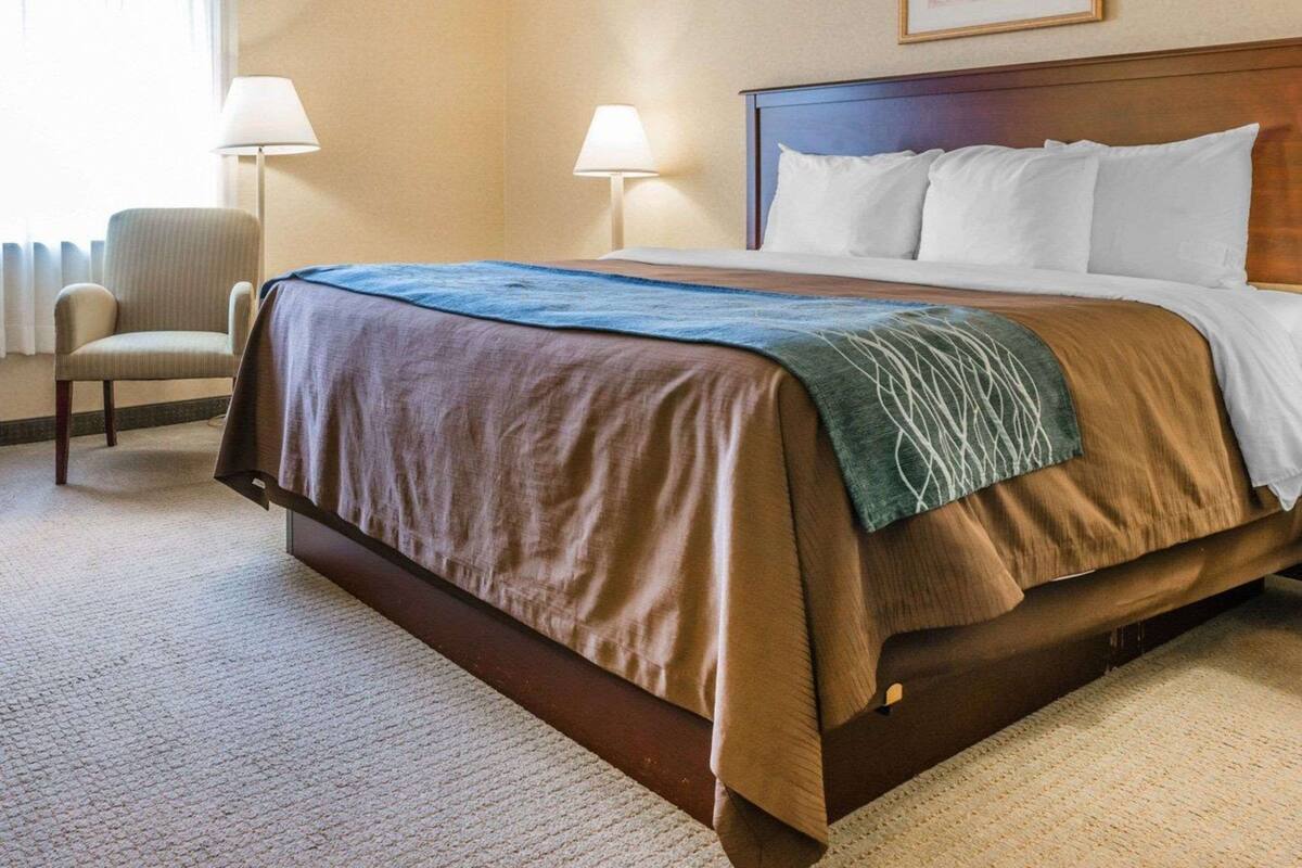 Quarto de hotel com cama, duas luminárias de cabeceira, uma cadeira e um quadro floral na parede.