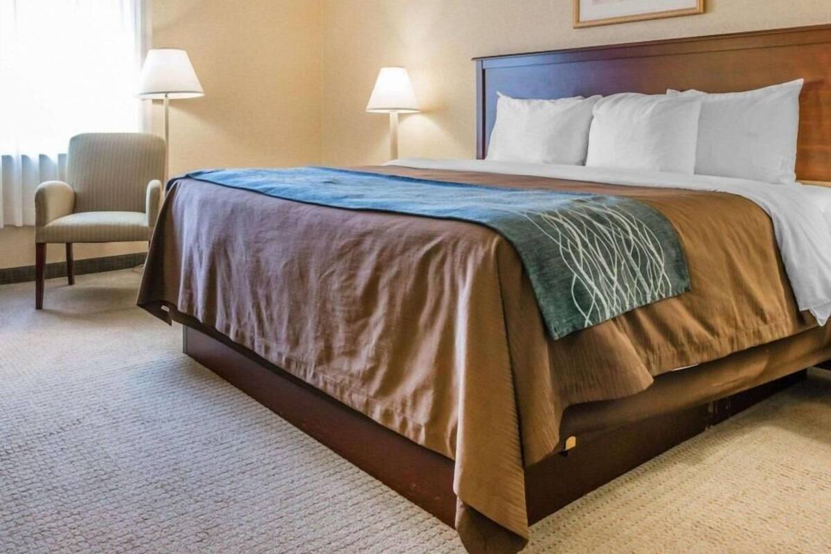 Quarto de hotel com uma cama, duas luminárias laterais, uma cadeira e um quadro na parede.