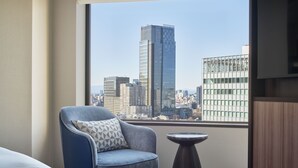 Quarto com vista para a cidade, uma poltrona azul com almofada geométrica, uma mesa redonda pequena e um armário de madeira.