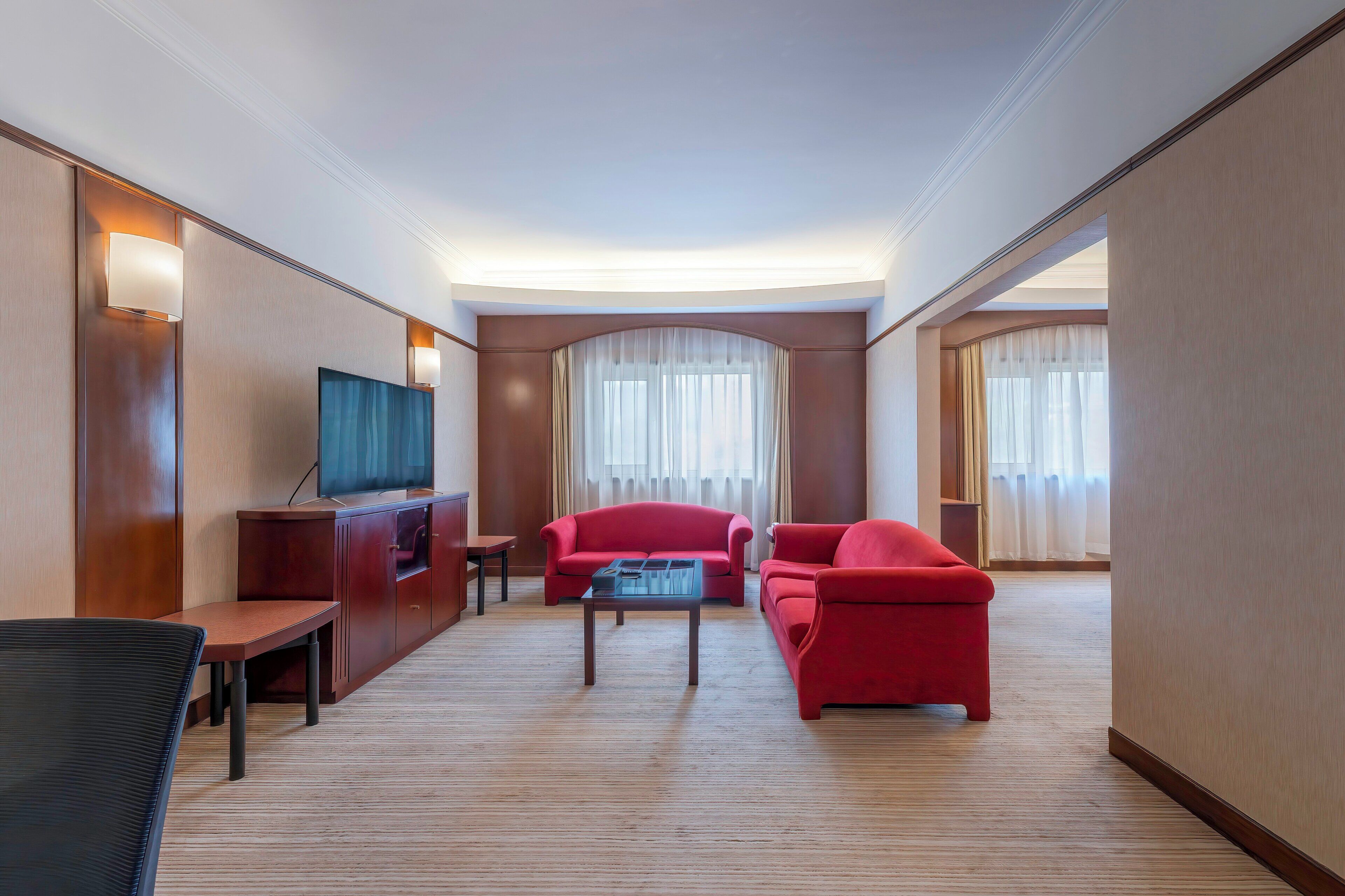 Quarto de hotel com televisão de tela plana, dois sofás vermelhos, uma mesa de centro de madeira e uma escrivaninha de madeira.