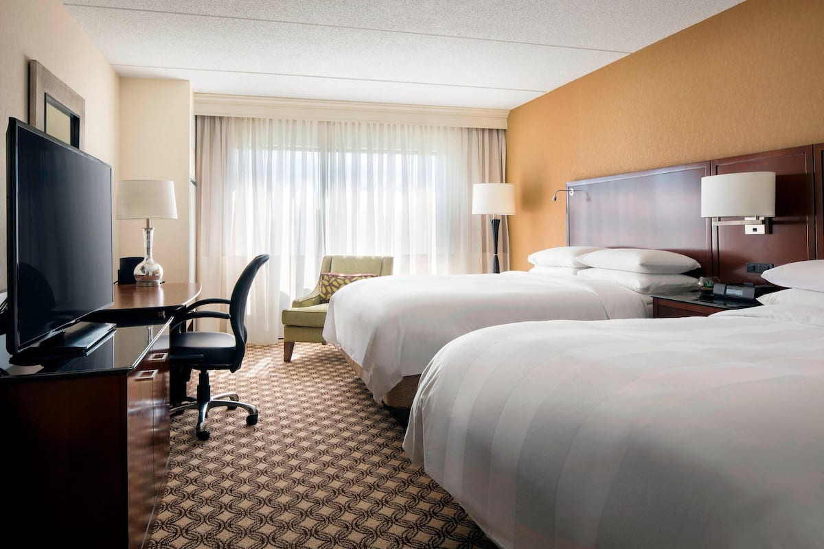 Quarto de hotel com duas camas, uma escrivaninha com televisão, uma cadeira e uma janela grande com cortinas.