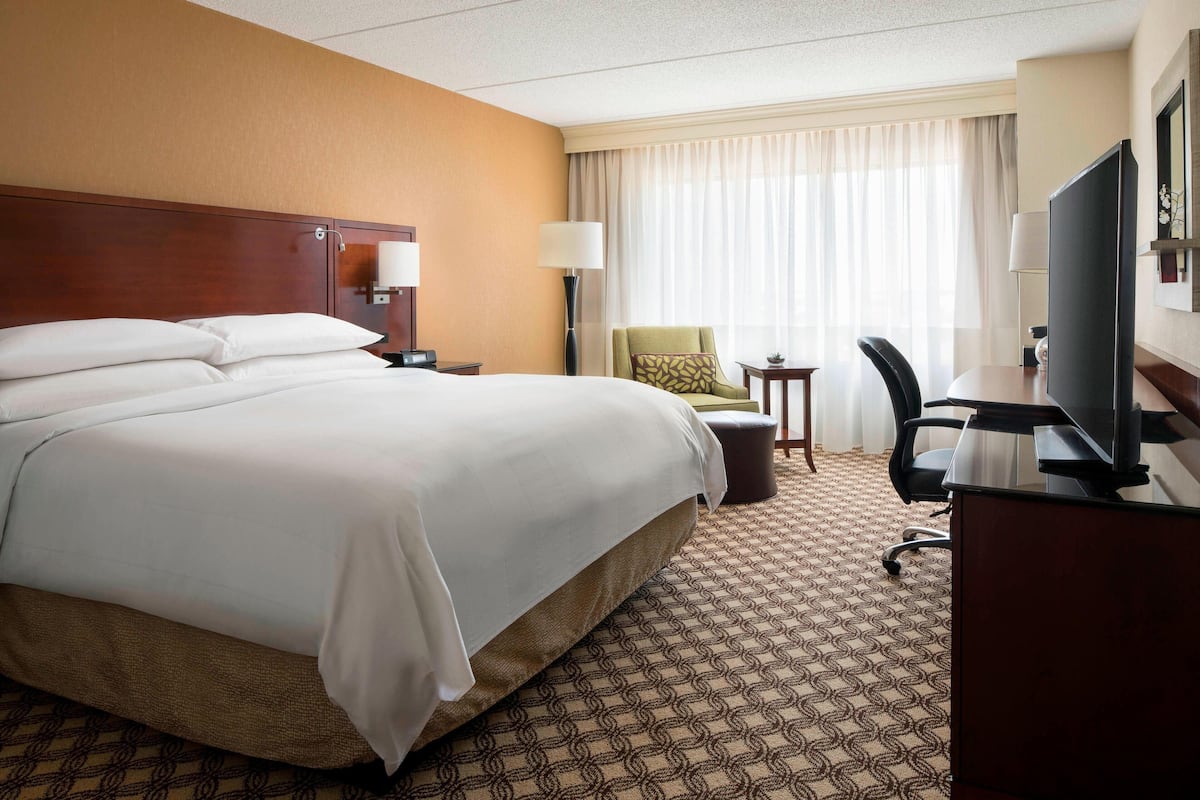 Quarto de hotel com uma cama grande, uma escrivaninha com computador, uma cadeira, um abajur e uma televisão.