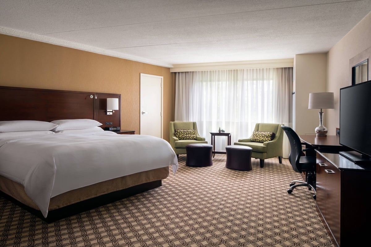 Quarto de hotel com uma cama grande, duas poltronas, uma escrivaninha com televisão e uma janela com cortinas.