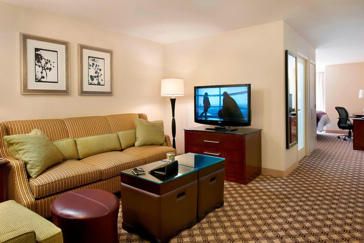 Quarto de hotel com sofá listrado, mesa de centro de vidro, televisão, abajur e tapete estampado.