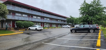 Um motel de vários andares com telhado vermelho e varandas. Vários carros estão estacionados no estacionamento, e uma árvore é visível à direita.