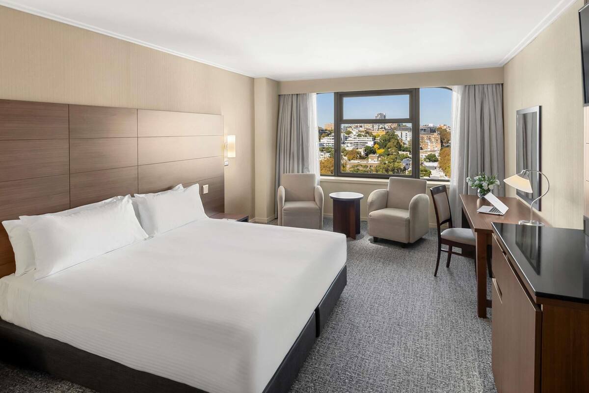 Quarto de hotel com uma cama grande, duas poltronas, uma escrivaninha e vista para a cidade.