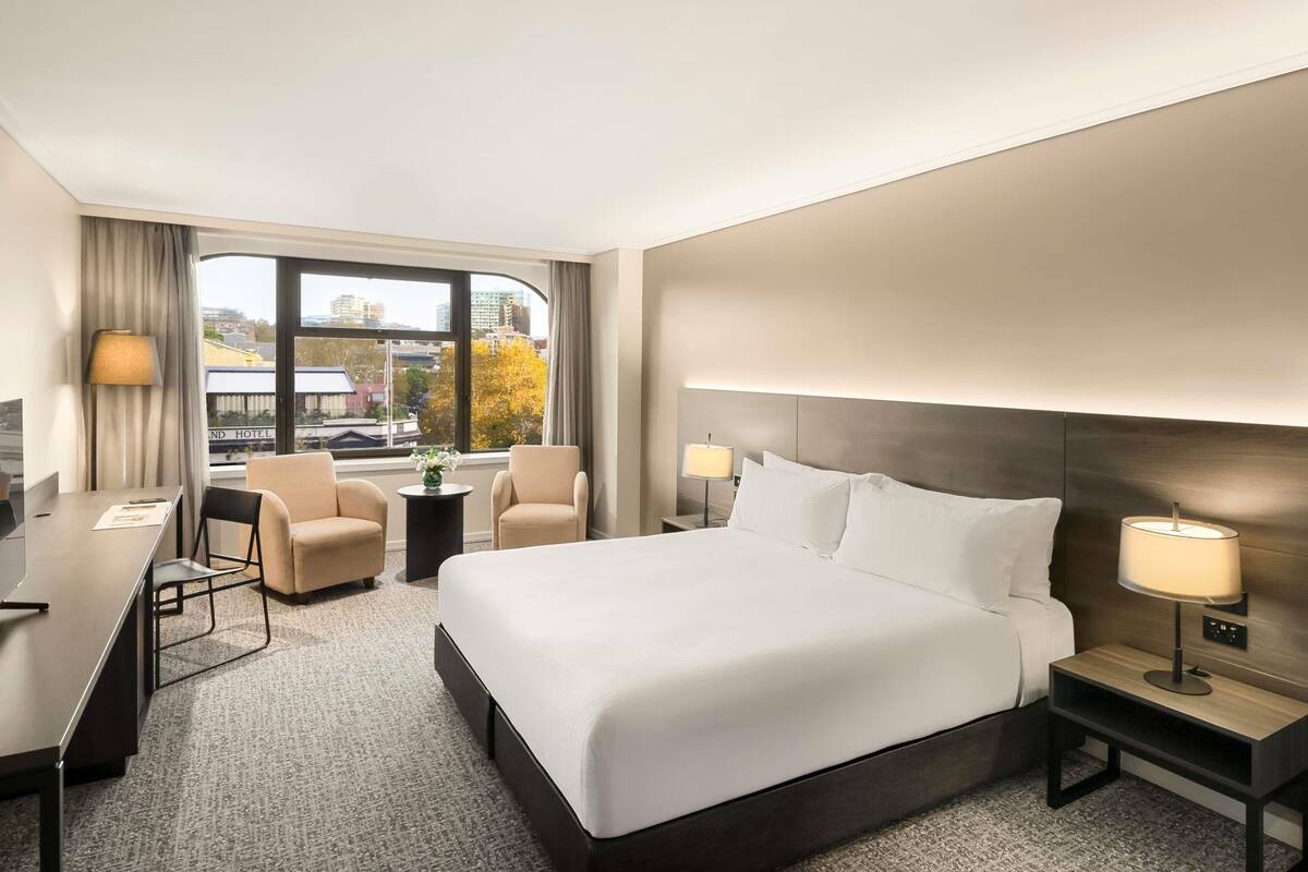 Quarto de hotel com uma cama grande, duas poltronas, uma escrivaninha e vista para a paisagem urbana.