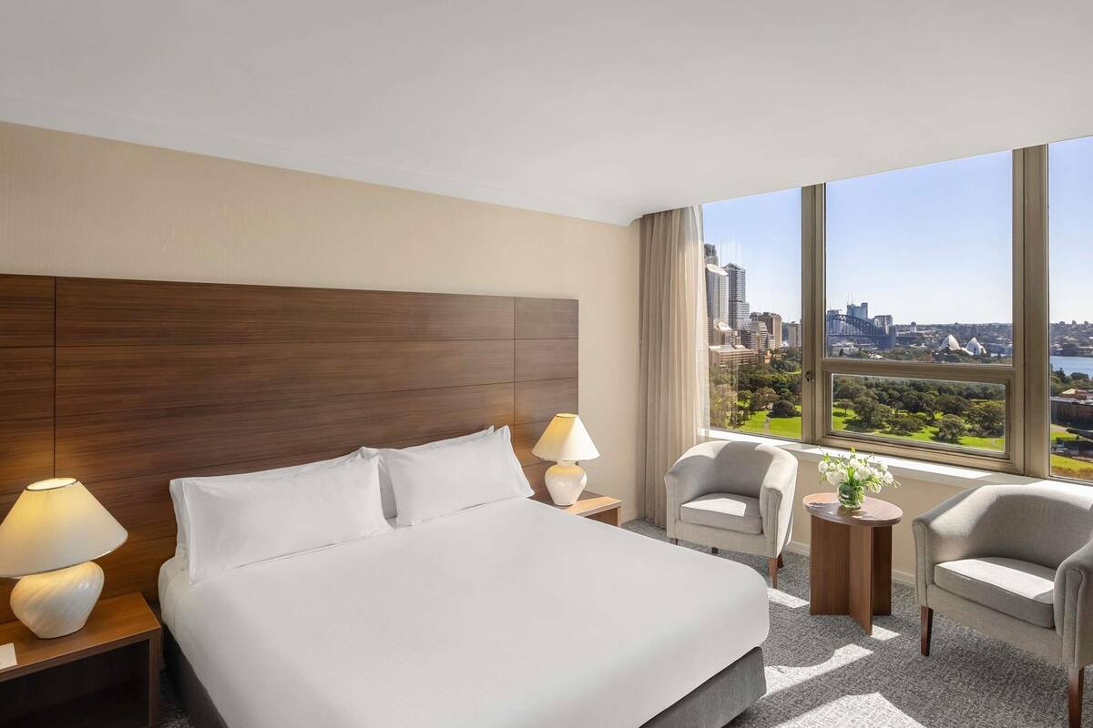 Quarto de hotel com uma cama grande, duas mesas de cabeceira com abajures, uma cadeira e vista para a cidade.