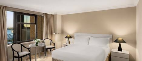 Quarto de hotel com uma cama grande, duas mesas de cabeceira, uma cadeira, uma mesinha redonda e vista para o mar.