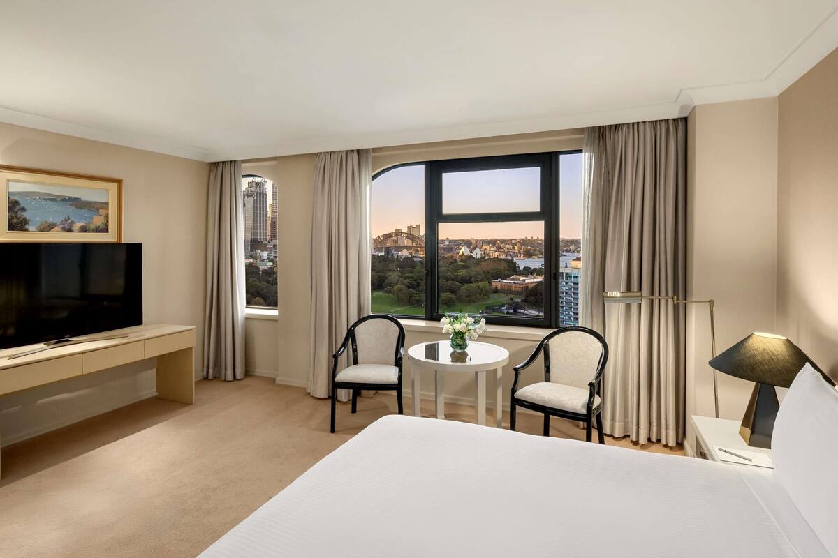 Quarto de hotel com cama, televisão, duas cadeiras, uma mesa pequena com um vaso e vista para a paisagem urbana através de amplas janelas.