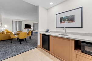 Quarto de hotel com cozinha compacta, área de jantar com cadeiras amarelas, televisão de tela plana e quadro na parede.