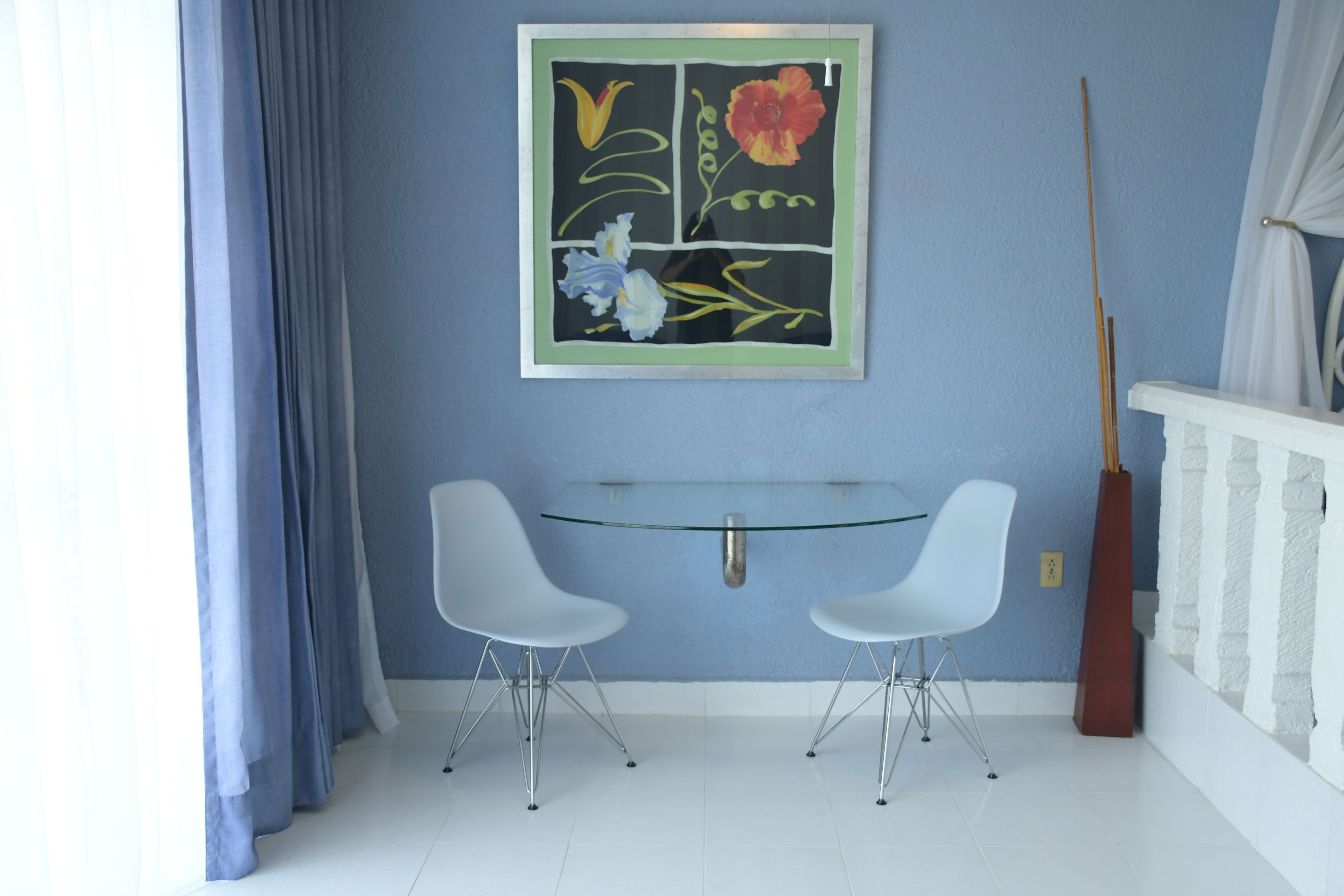 Um quarto com mesa de vidro, duas cadeiras brancas, uma pintura floral emoldurada, um objeto de madeira alto e uma cortina azul.