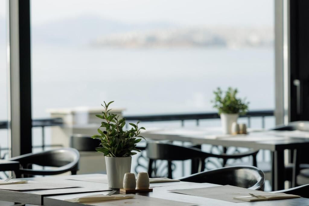 Restaurante com mesas montadas para refeições, plantas em vasos e vista para a água.