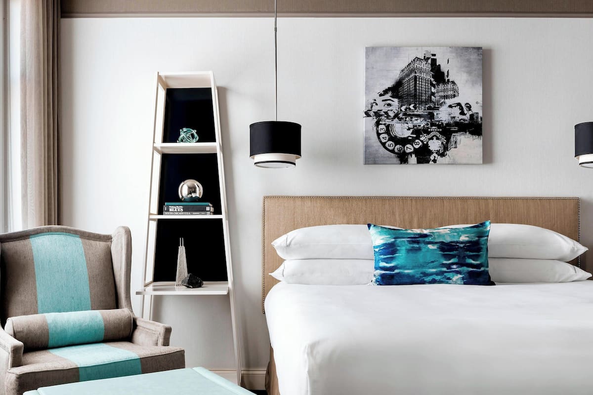 Quarto de hotel com cama, uma cadeira, prateleira com objetos decorativos e uma obra de arte na parede.