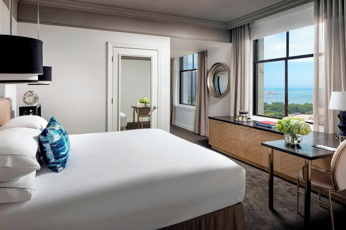 Quarto de hotel com uma cama grande, uma escrivaninha com abajur, vista para a cidade e uma janela com cortinas.