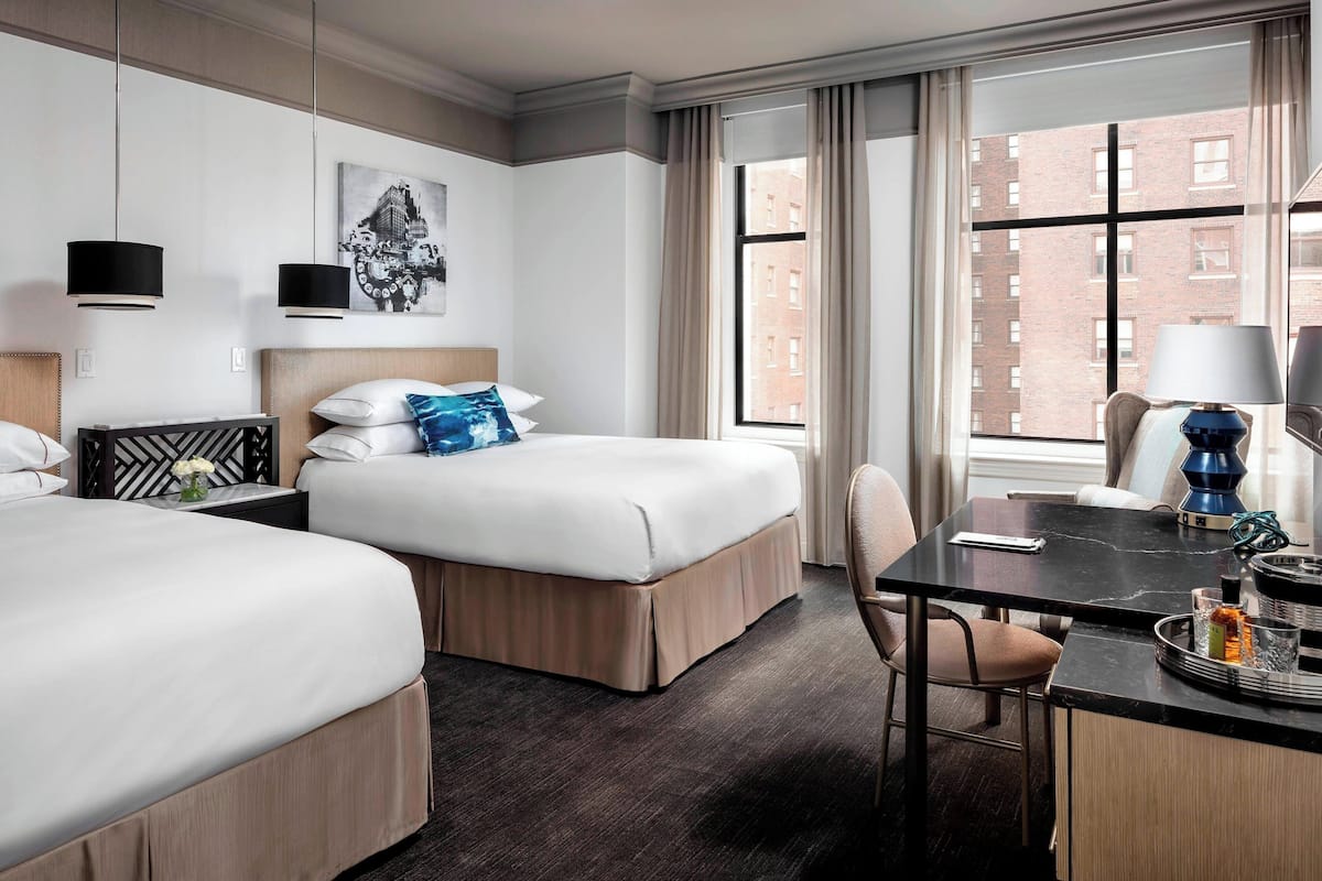 Quarto de hotel com duas camas, uma mesa de jantar, uma cadeira, um abajur e vista para a cidade através de amplas janelas.