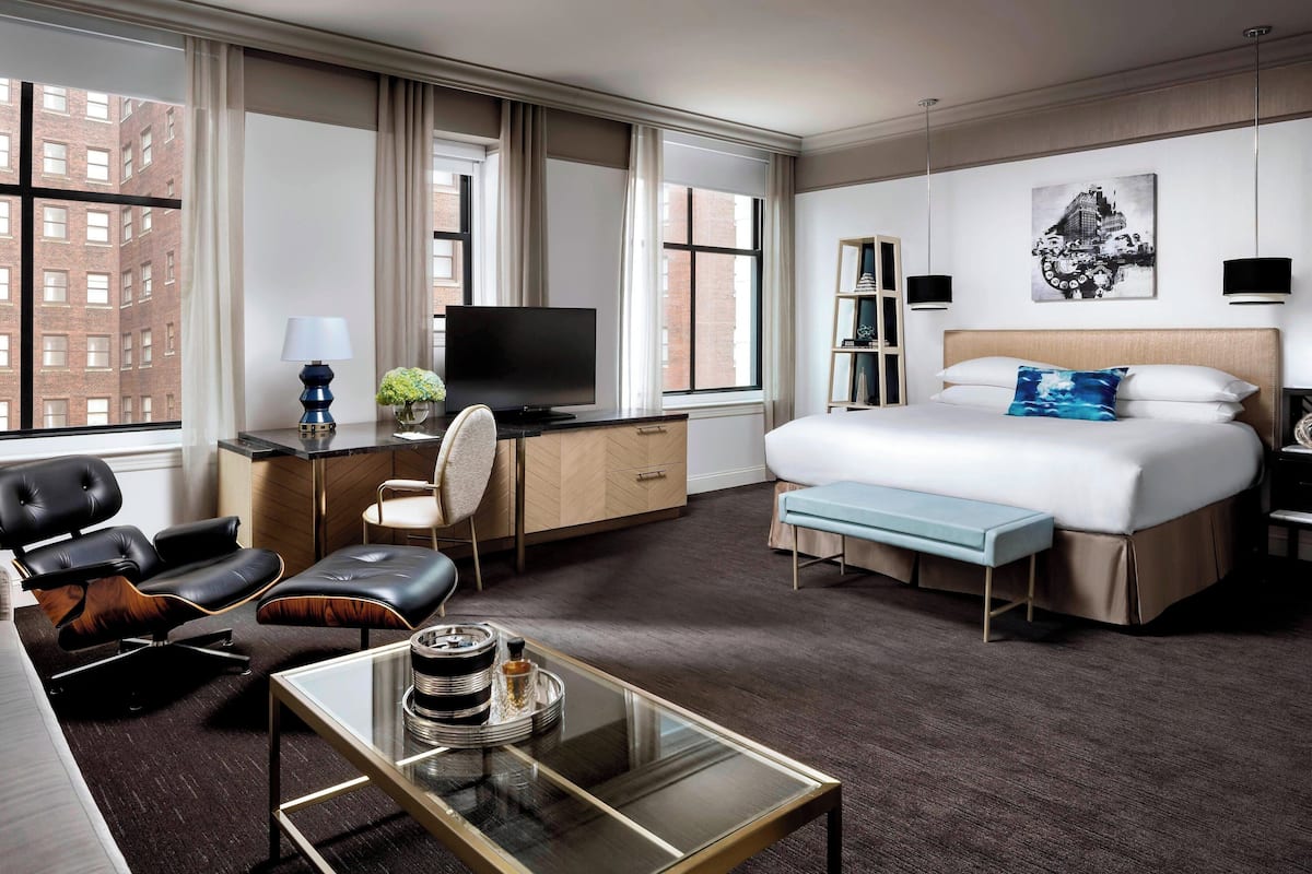 Quarto de hotel moderno com uma cama grande, uma escrivaninha com televisão, um sofá de couro e uma mesa de centro de vidro.